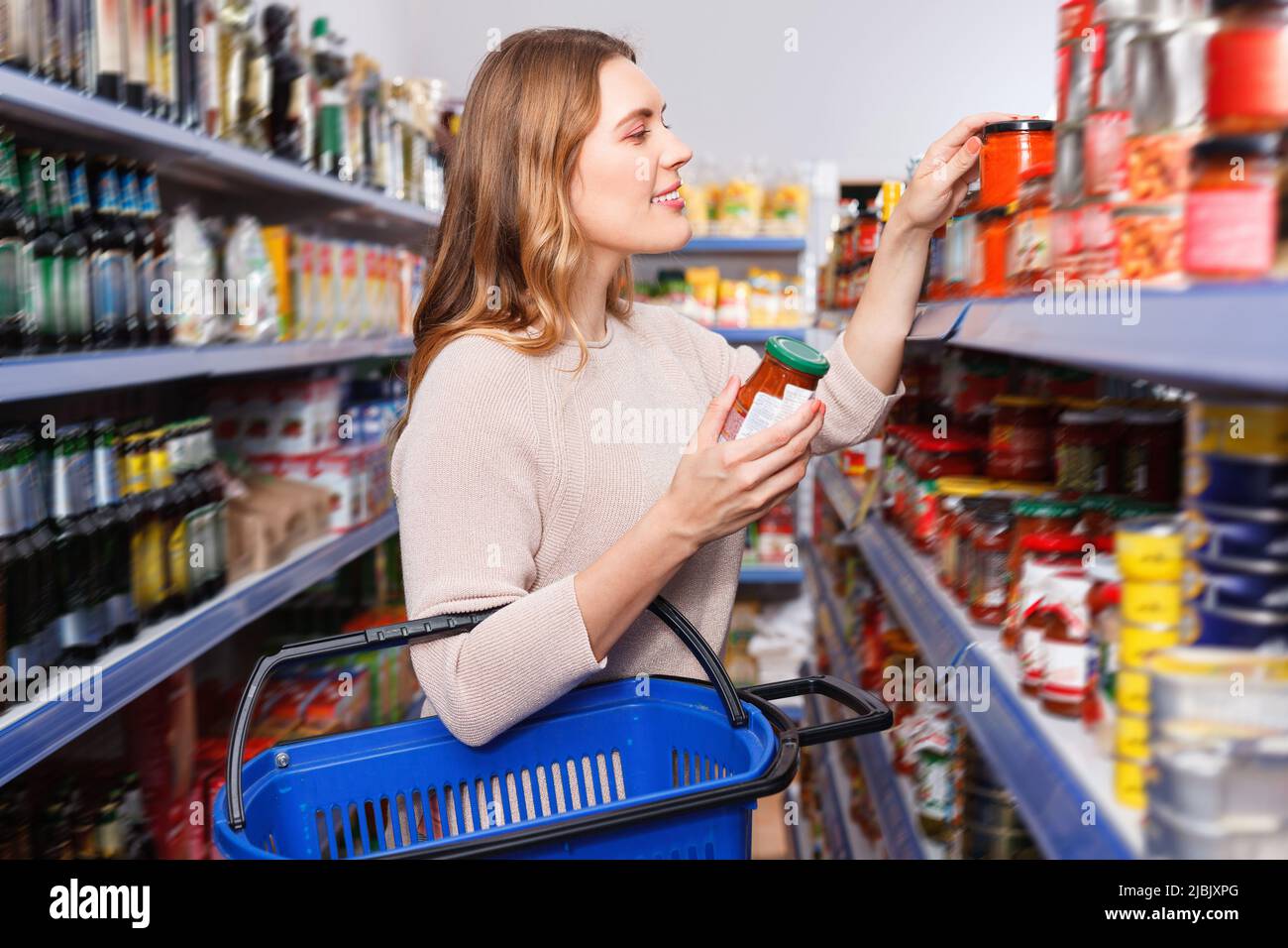 Positive female customer with basket holding canned goods in the food ...