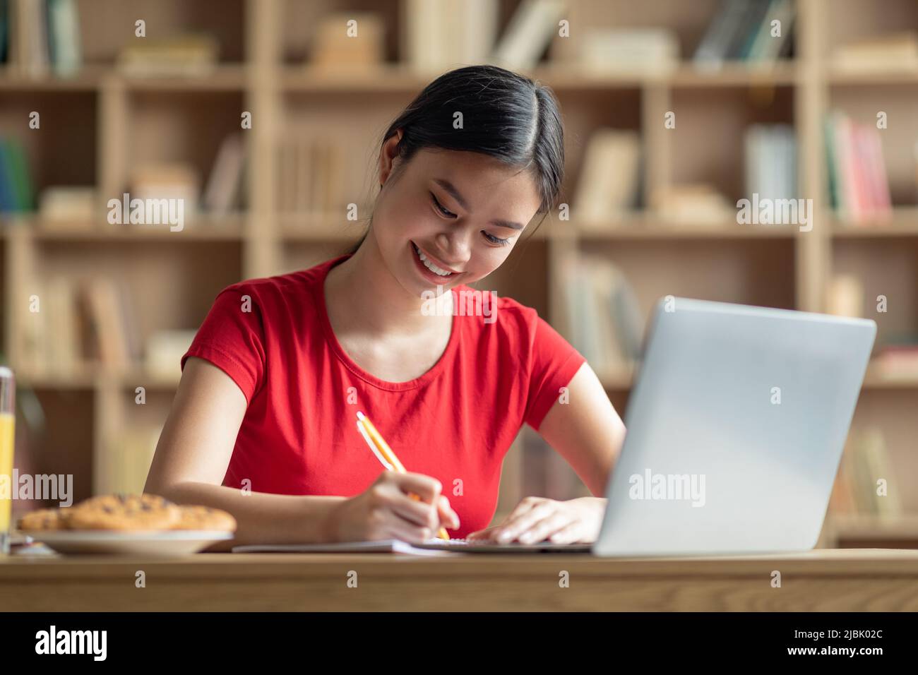 Glad teenager asian female makes notes at table with laptop, study in ...