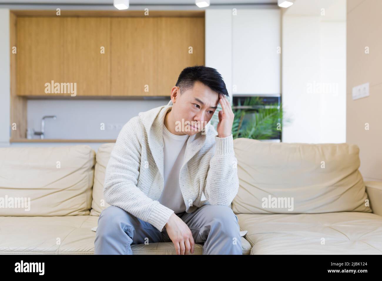 young asian man sitting on sofa at home pensive, worried about problems ...