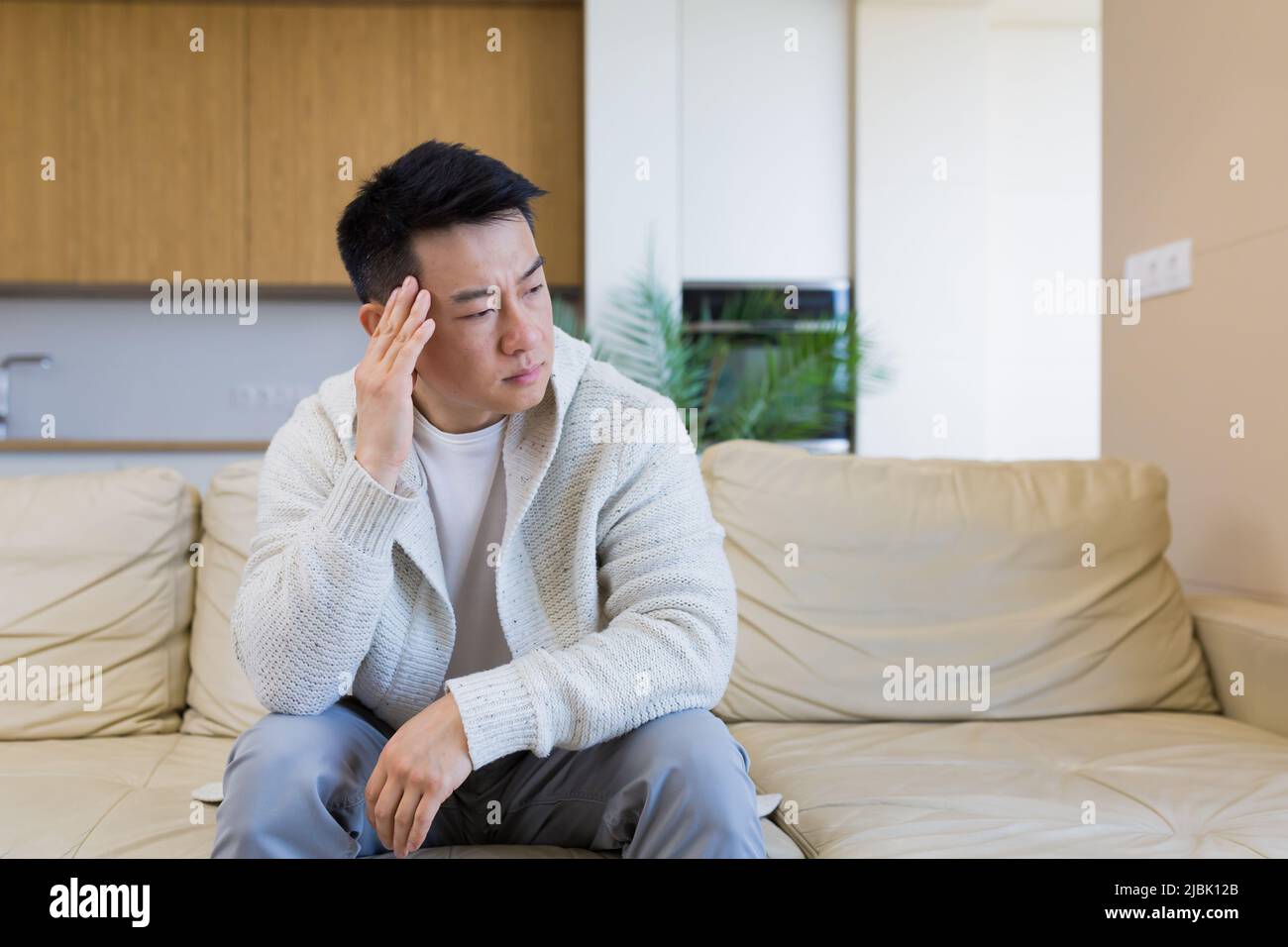 young asian man sitting on sofa at home pensive, worried about problems ...