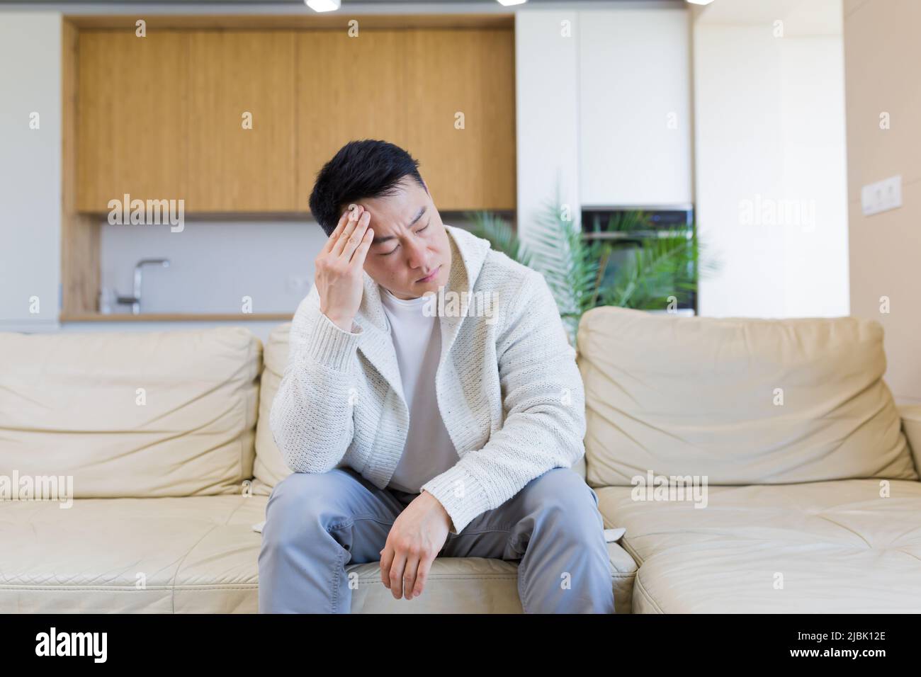 young asian man sitting on sofa at home pensive, worried about problems ...