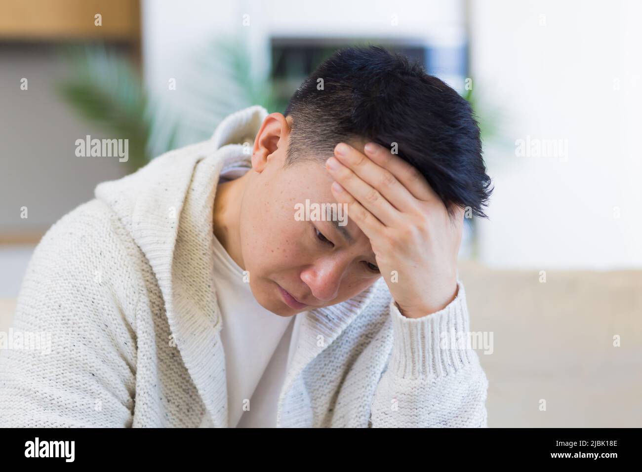 young asian man sitting on sofa at home pensive, worried about problems ...