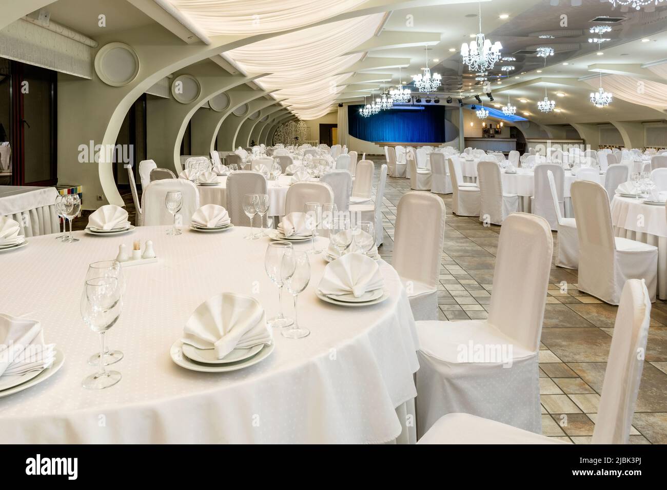 Banquet hall decorated in white with round served tables and stage for ...
