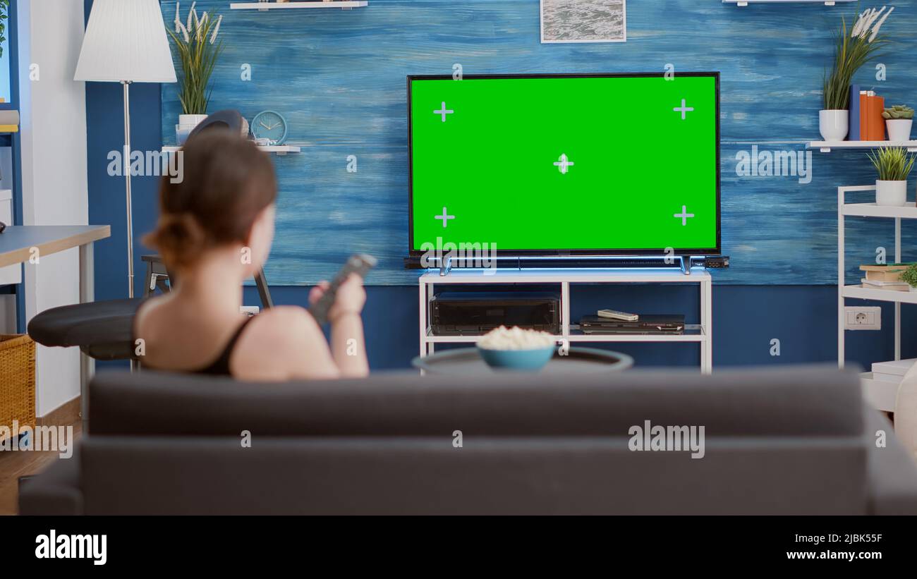 Young woman sitting on sofa looking at green screen on tv and switching ...