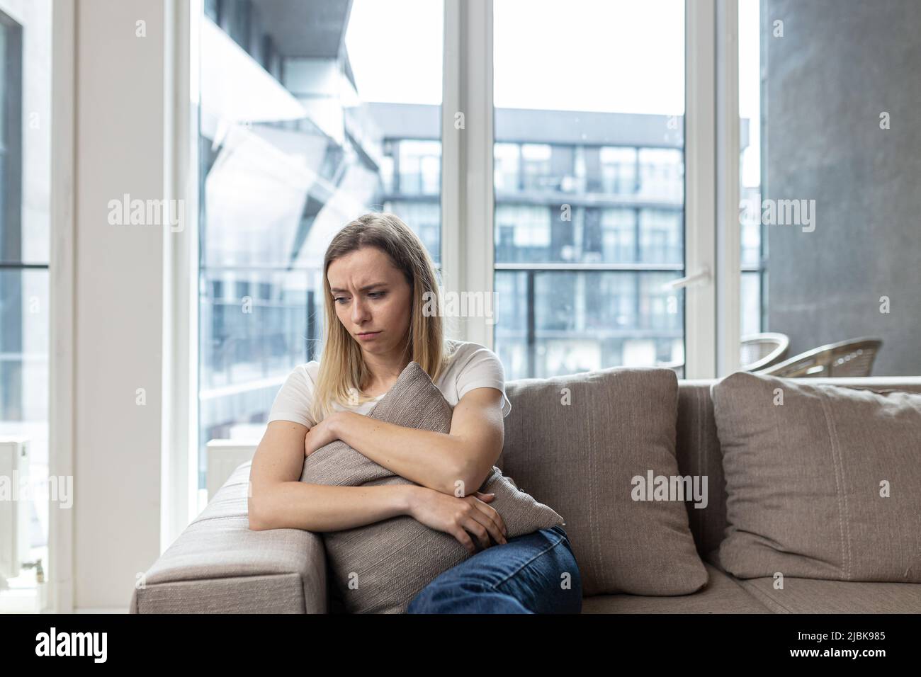 Upset young woman sitting on sofa at home, hugging pillow, feeling bad ...