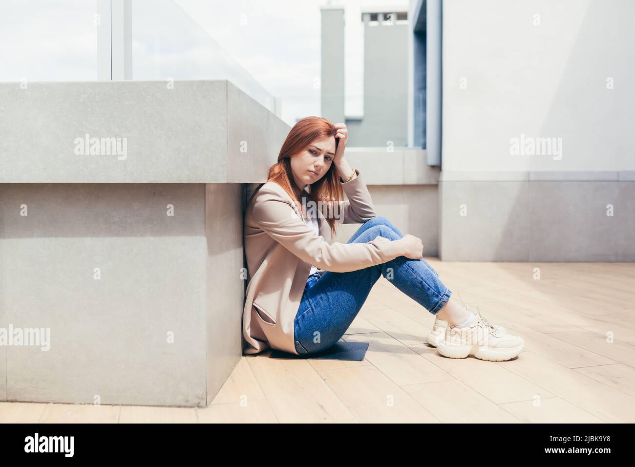 Sad female office worker sitting on the balcony floor of a modern ...