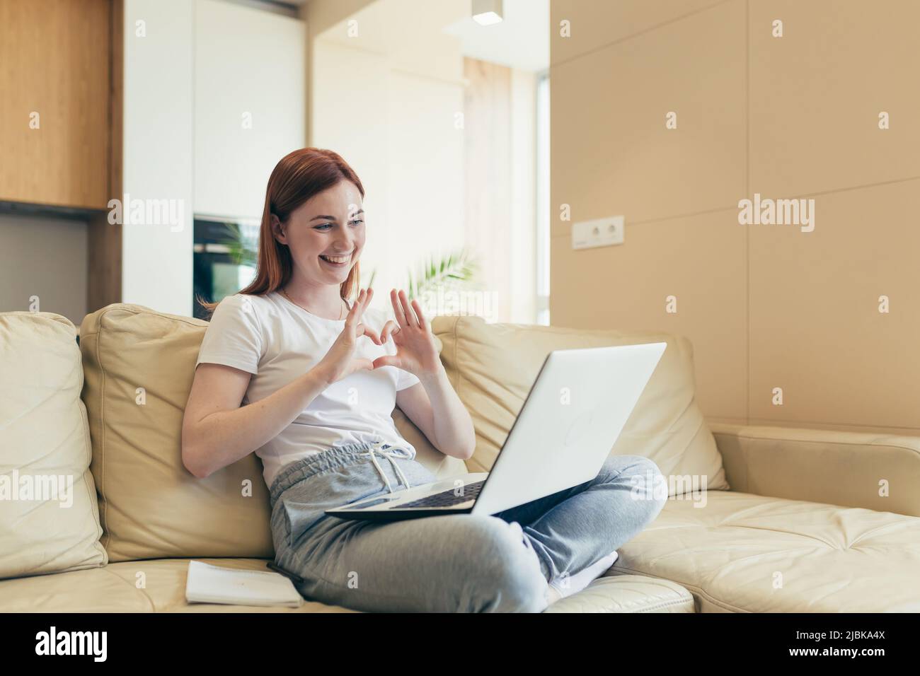 young happy girl talking on video call online with laptop webcam at home showing heart gesture ...
