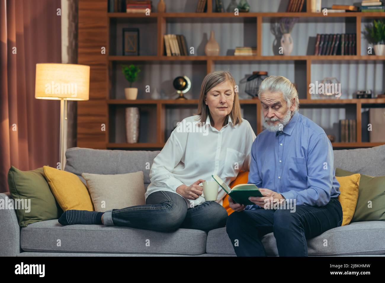 senior couple spending leisure time reading a book together sitting on cozy sofa at home ...
