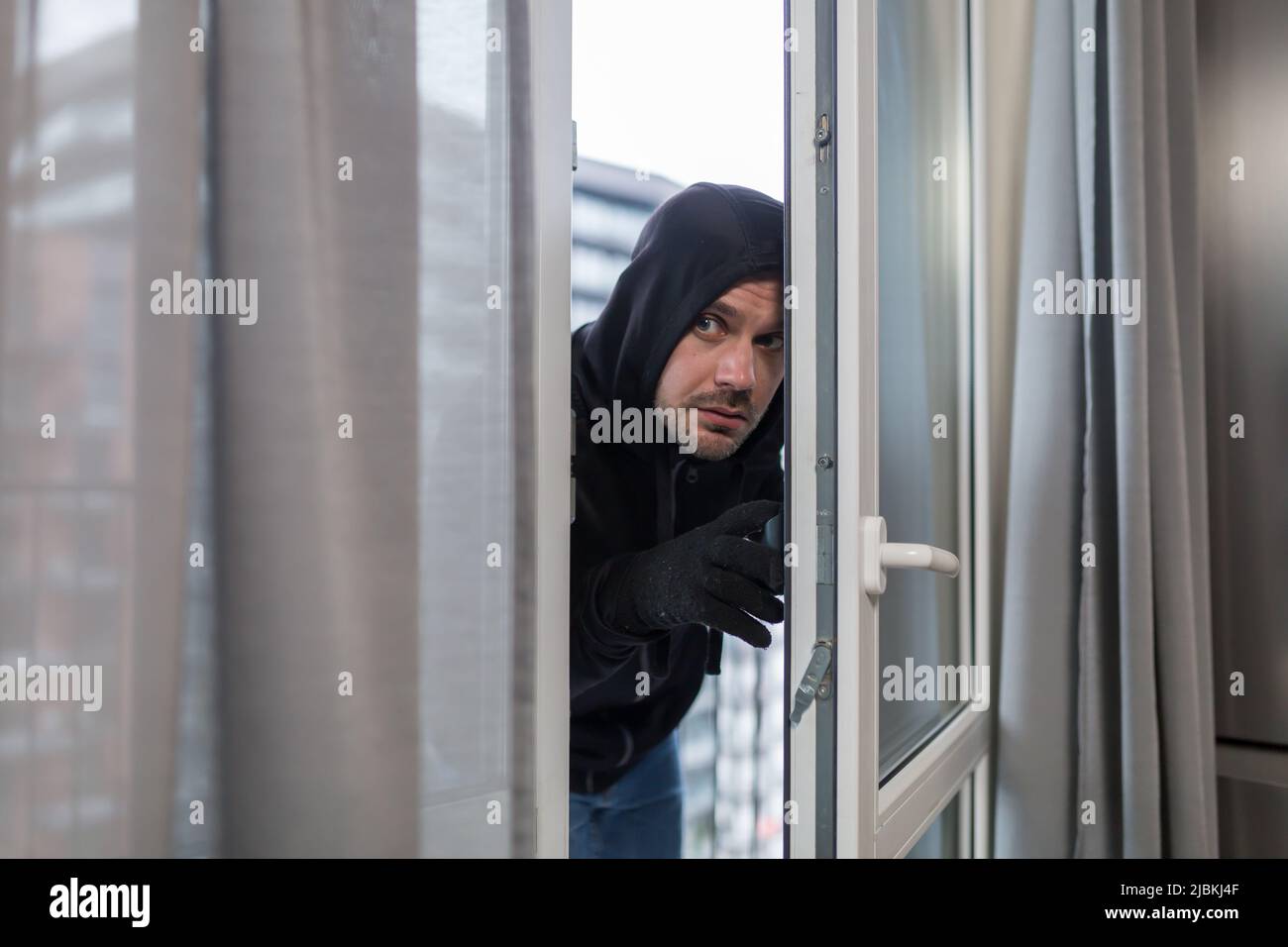 Young male robber in dark clothes looks out the window of the apartment ...