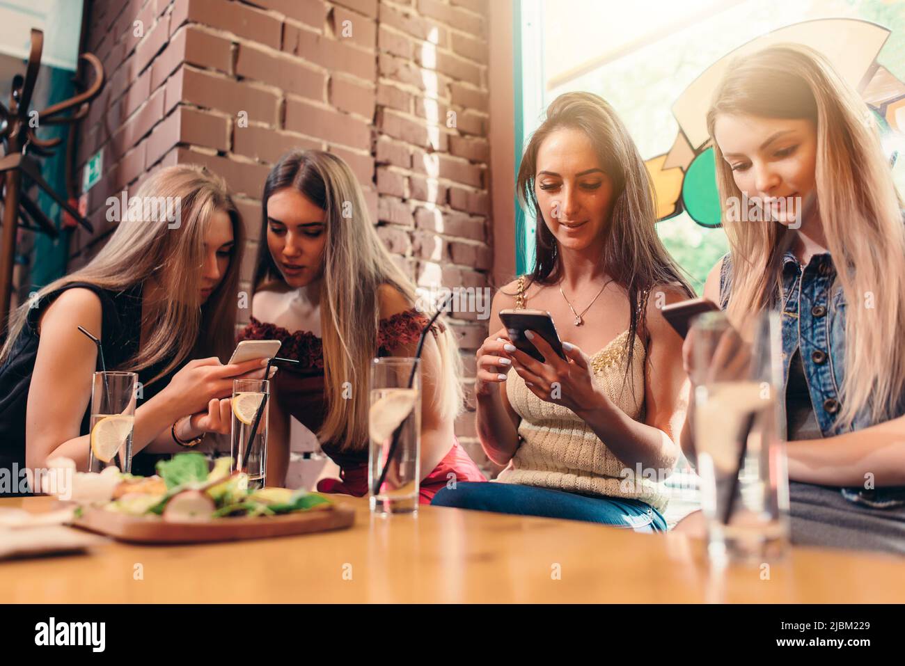 Four smiling female students sitting in cafeteria chatting using mobile ...