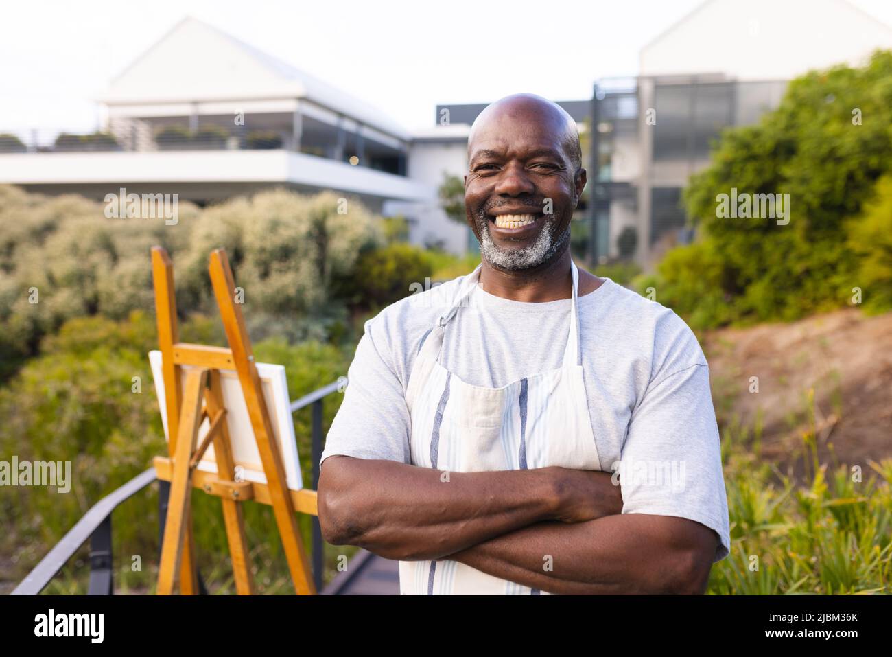 Portrait of smiling bald african american senior man with arms crossed standing by canvas and ...