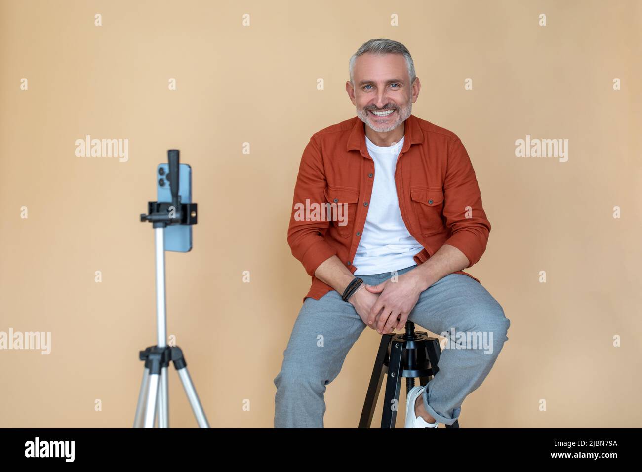 A man in a red jacket sitting on a stool in a studio and making selfie ...