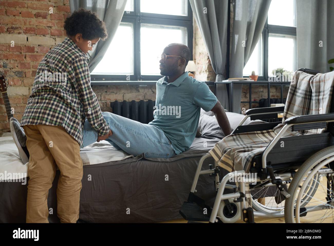 Little son holding legs of his disabled father to help him lie on bed ...