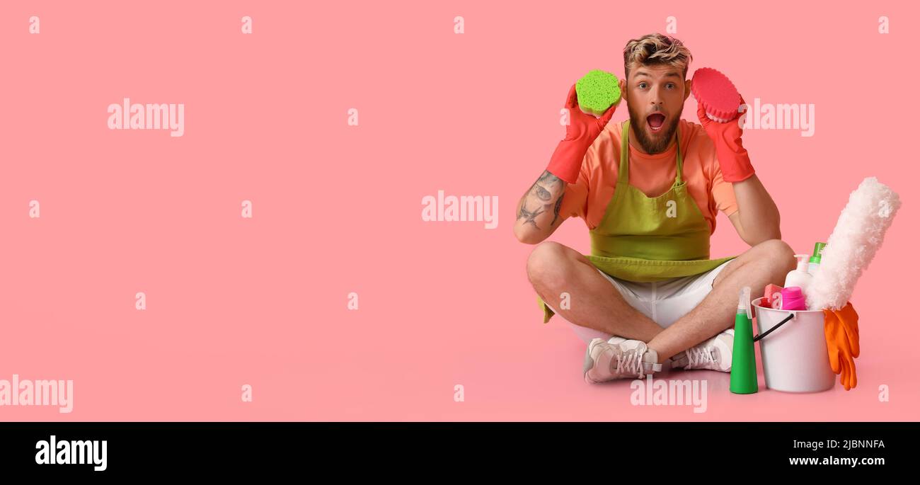 Surprised young man with cleaning supplies sitting on pink background ...