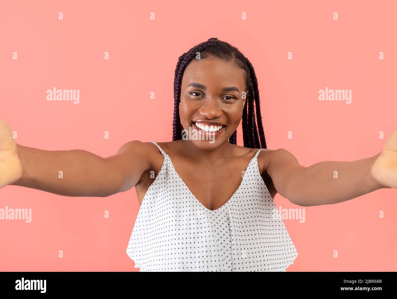 Smiling young black woman taking selfie with smartphone, chatting online on pink studio ...