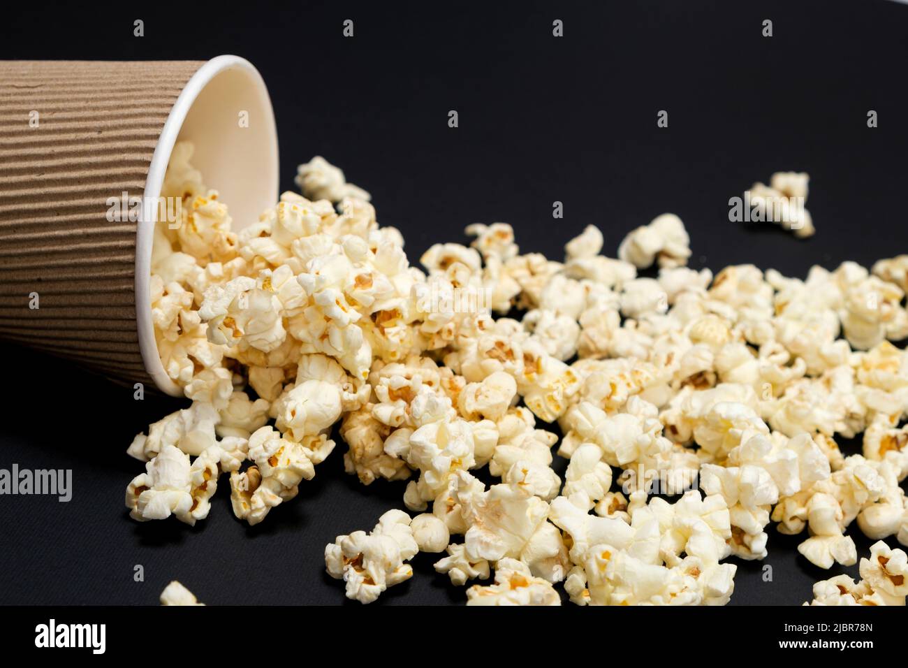 Popcorn spills out of a brown paper cup on a black background close-up ...