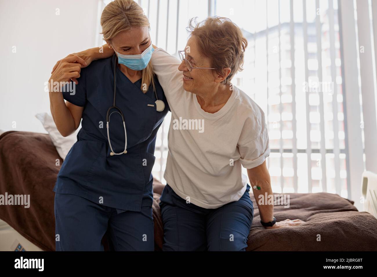 Female doctor in mask helping patient to get up from bed in the ward at ...