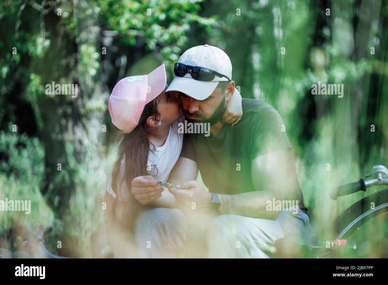 Portrait of family wearing caps sitting in park around trees after riding bike. Little girl ...