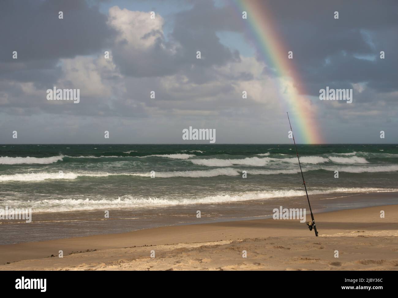 Surf caster fishing rod secured in the sand and rainbow in stormy sky ...