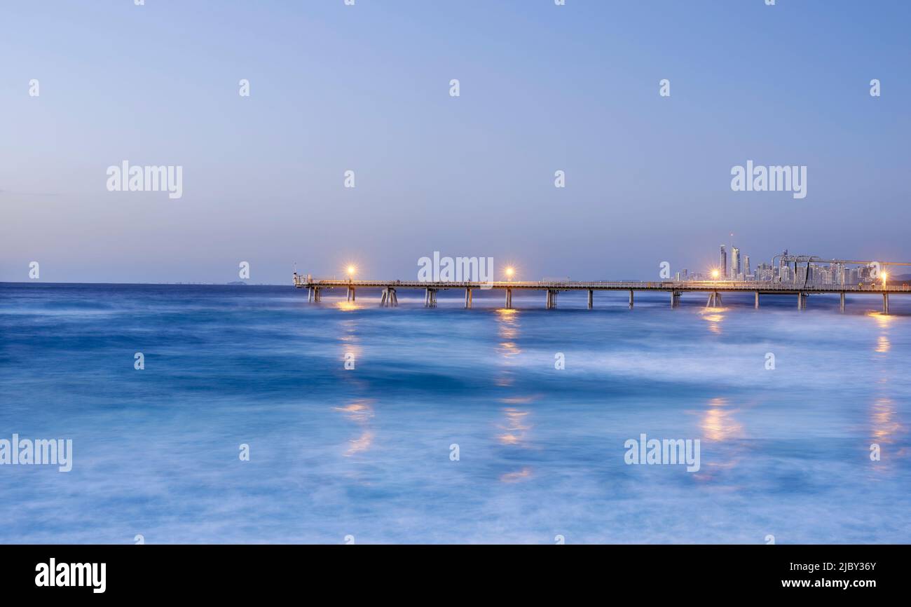 Long pier stretching out into calm ocean in early evening with lights ...