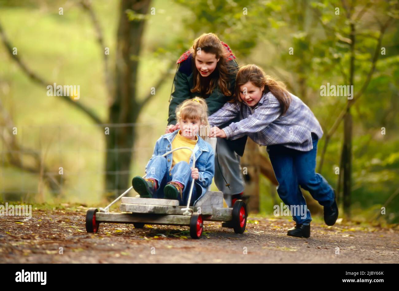 Girls pushing younger girl on trolley through park Stock Photo - Alamy