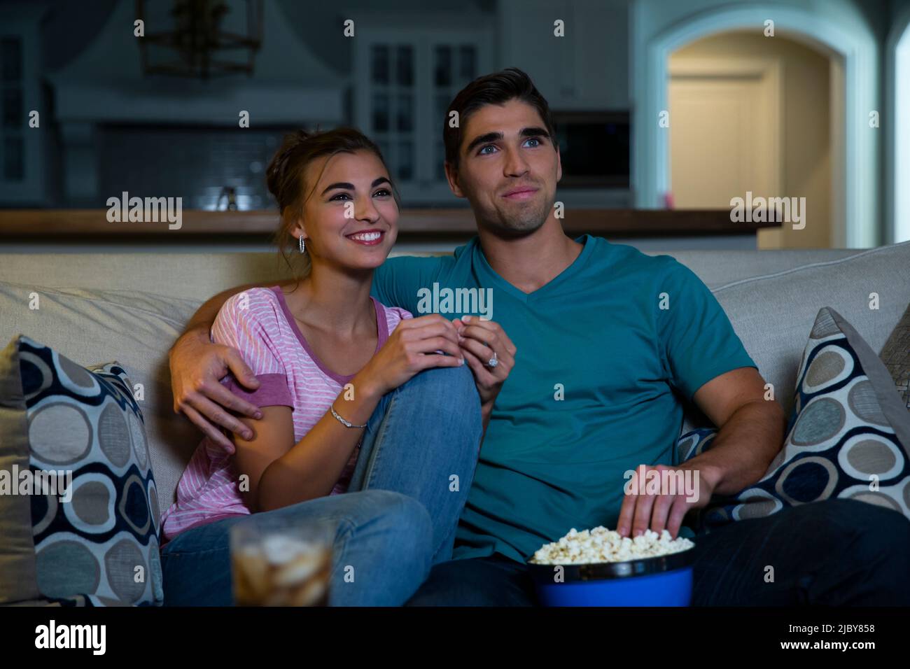 Young couple sitting on couch watching a movie at home with a bowl of ...
