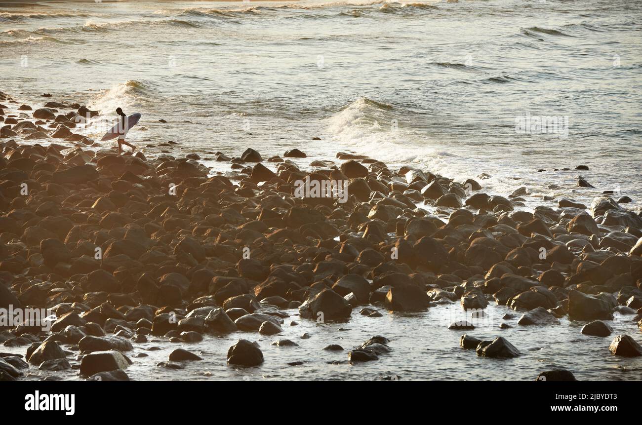 Lone Surfer carrying surfboard walking over rocks from the sea Stock ...