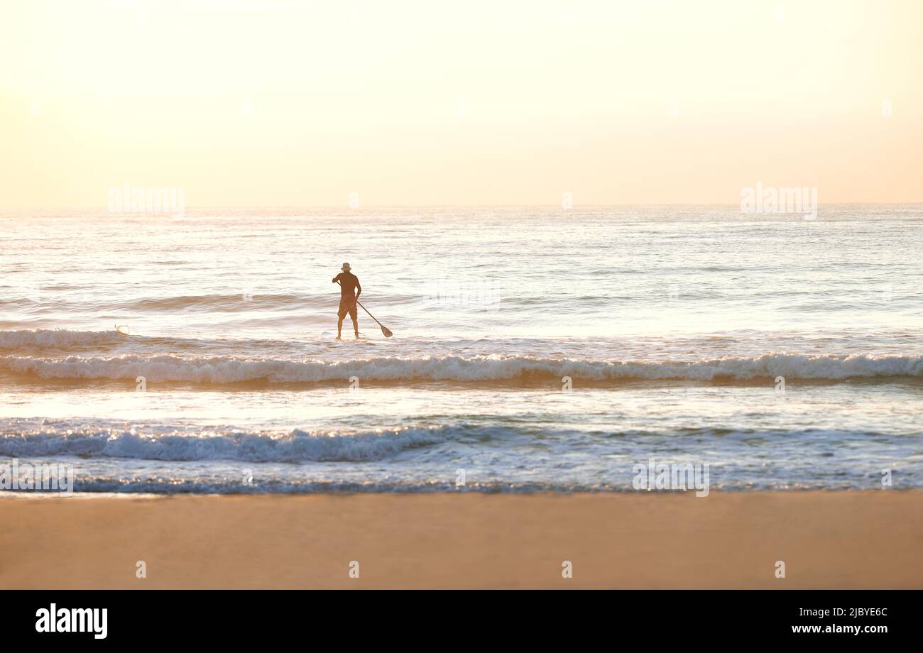 Back view of man paddle boarding through gentle waves at sunrise at the ...