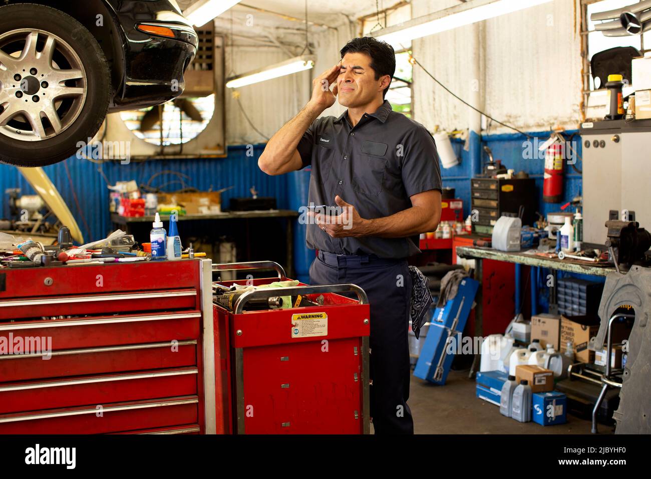 Hispanic car mechanic working in auto shop, having migraine headache ...