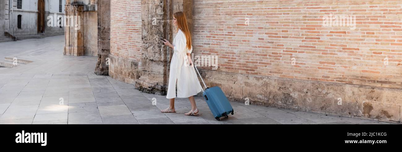 full length of redhead woman walking with luggage and using smartphone ...