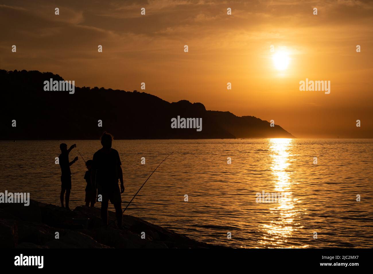 fishermen in silhouette at sunset on the sea cliff of Pesaro in Italy ...