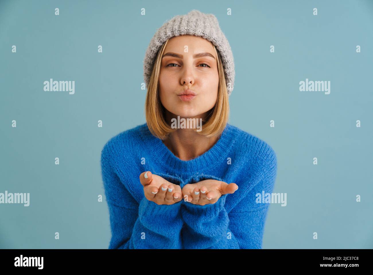 Young blonde woman wearing knit hat blowing air kiss isolated over blue ...
