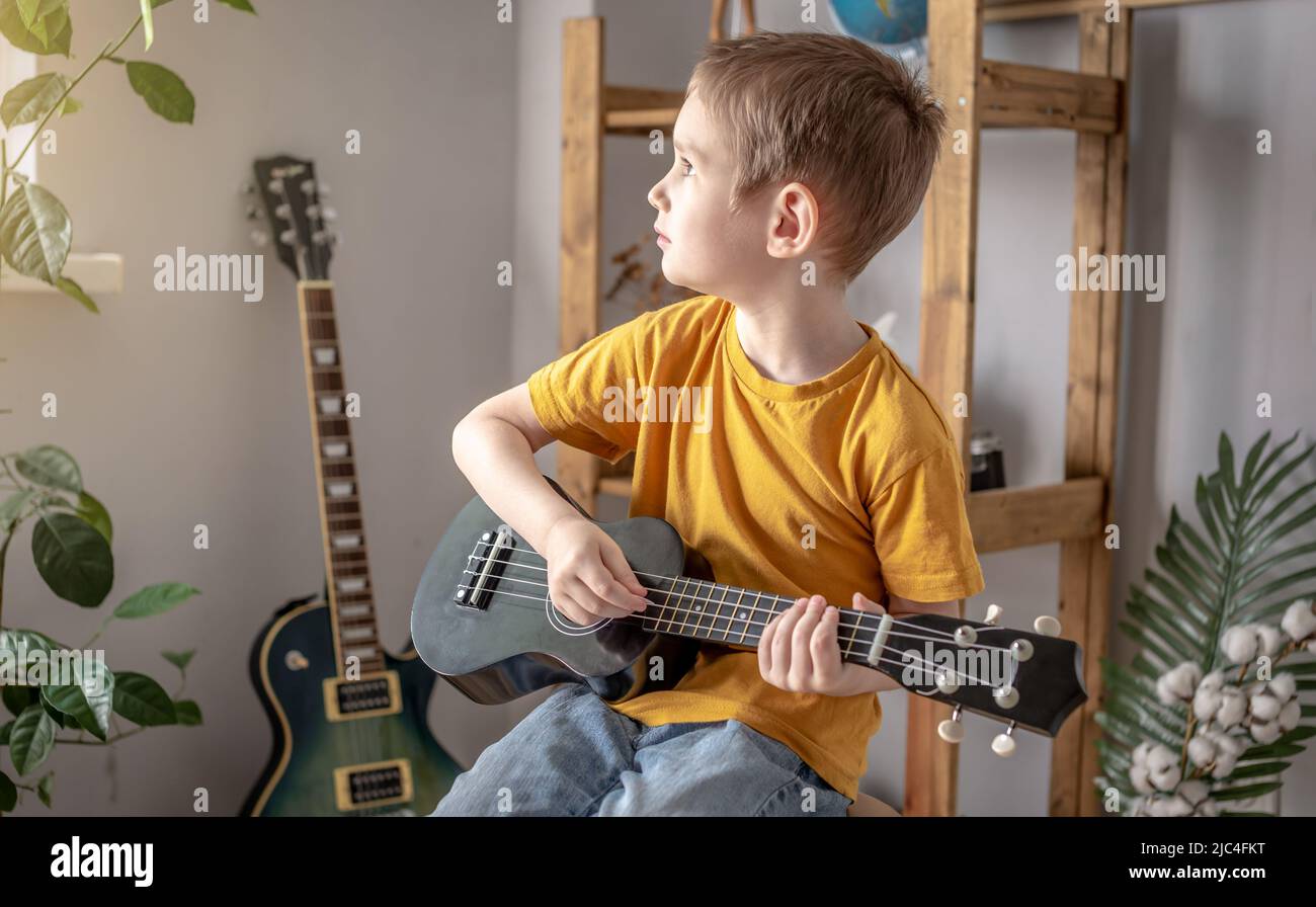 Cute funny boy is playing ukulele guitar in the music room. Joyful ...