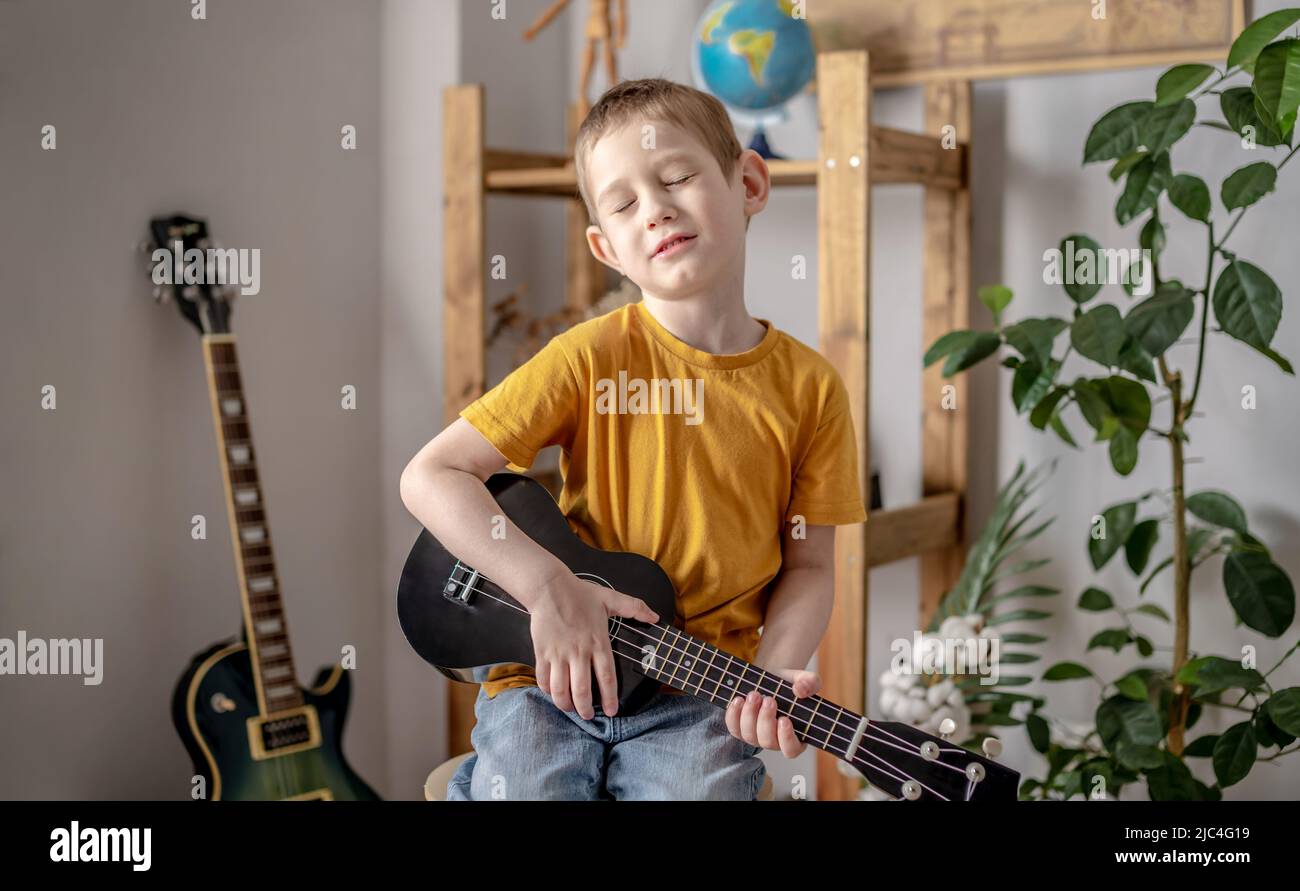 Cute funny boy is playing ukulele guitar in the music room. Joyful ...