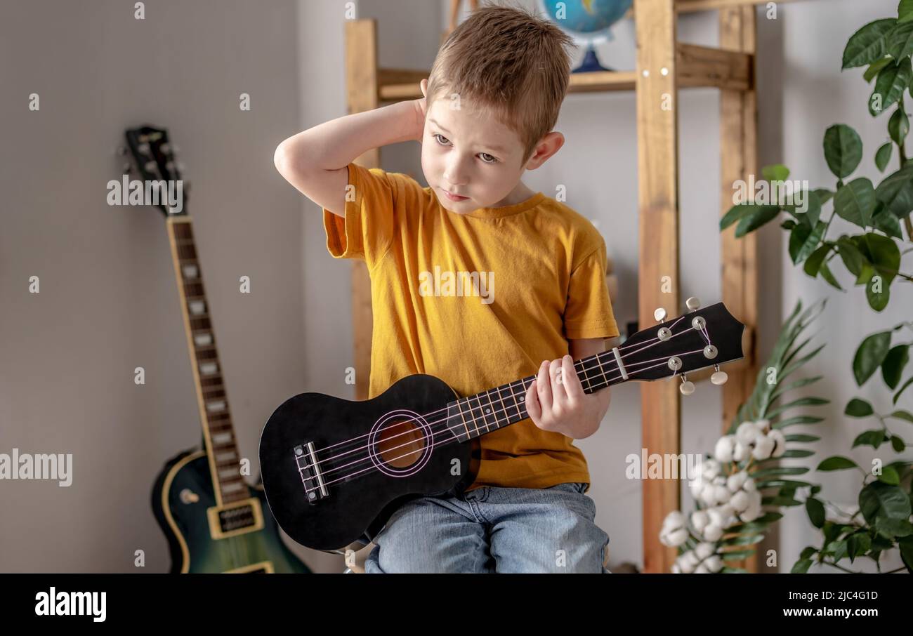 Cute funny boy is playing ukulele guitar in the music room. Joyful ...