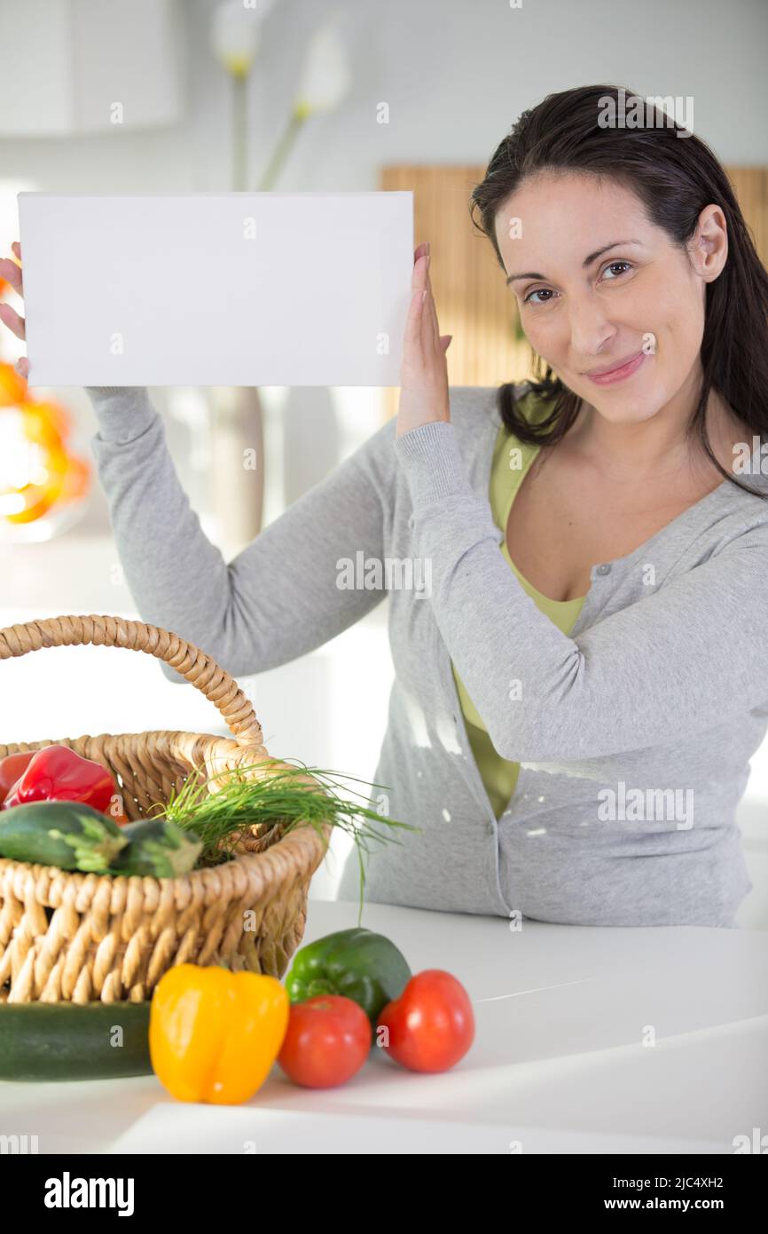 woman in kitchen with basket of vegetables holding blank sign Stock ...