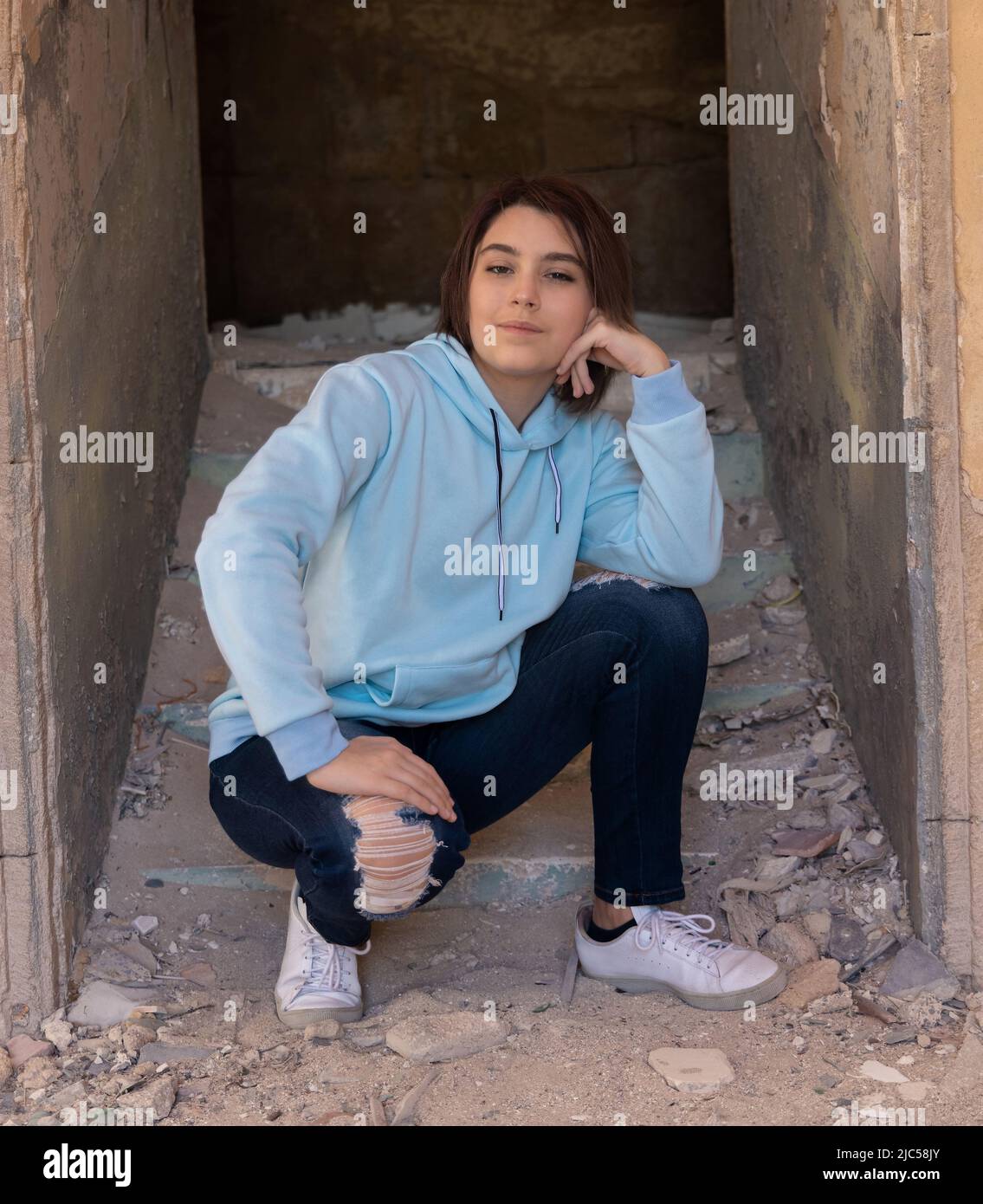 Teenager girl in light blue hoodie sitting on her haunches near a wall ...