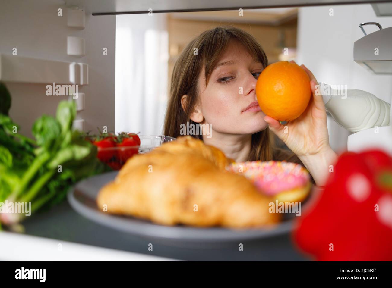 Woman sniffing a fresh orange and looking at other foods in the fridge ...