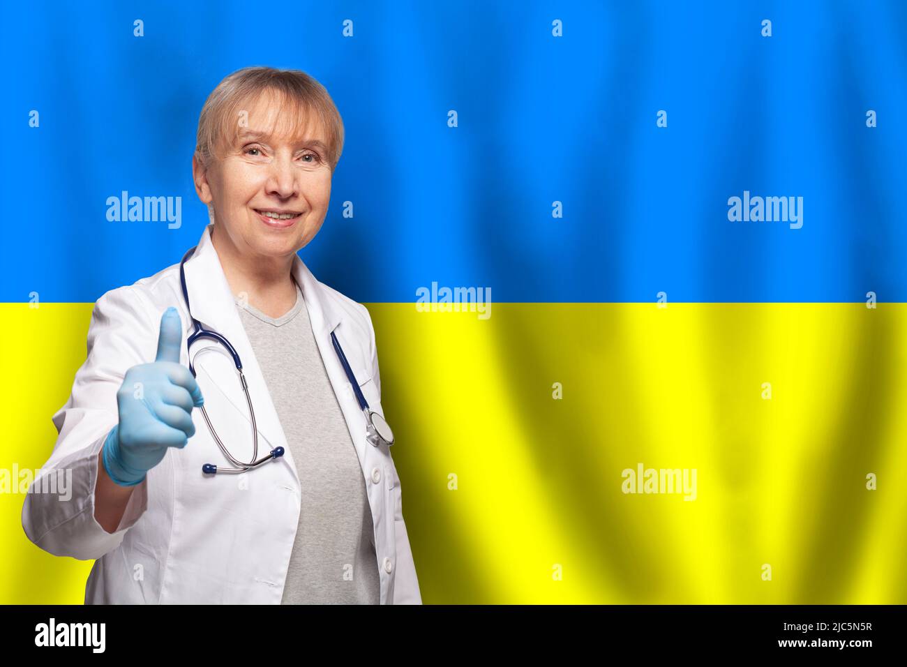 Smiling mature doctor woman holding stethoscope on flag of Ukraine ...