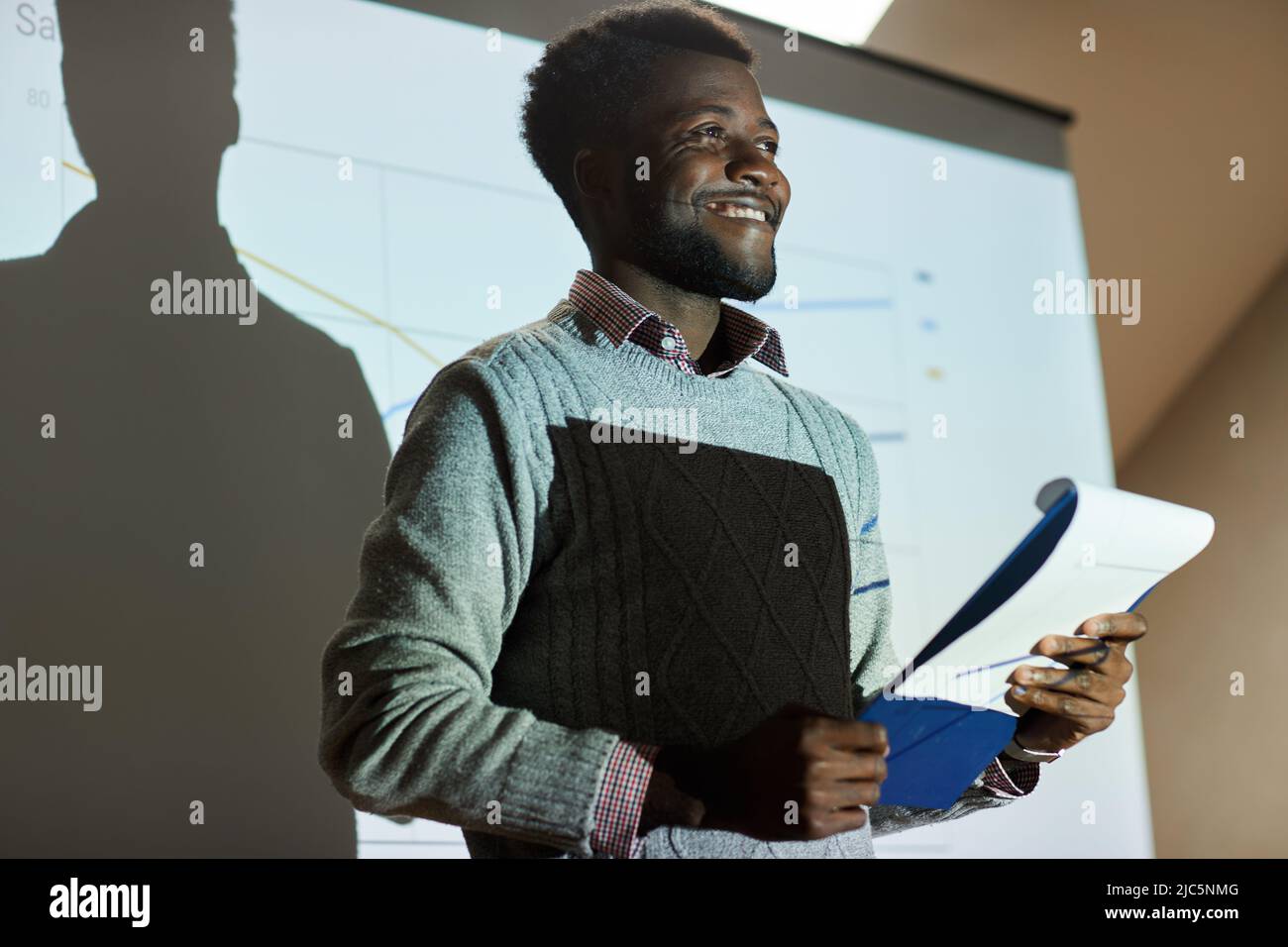 Smiling successful Black student with beard standing against projection ...