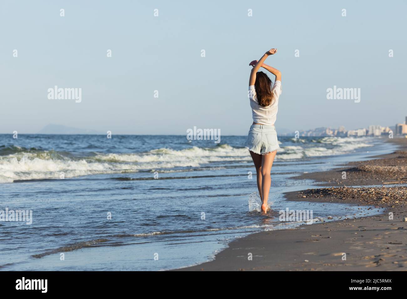 Back view of woman in denim shorts and shirt walking near sea on beach ...