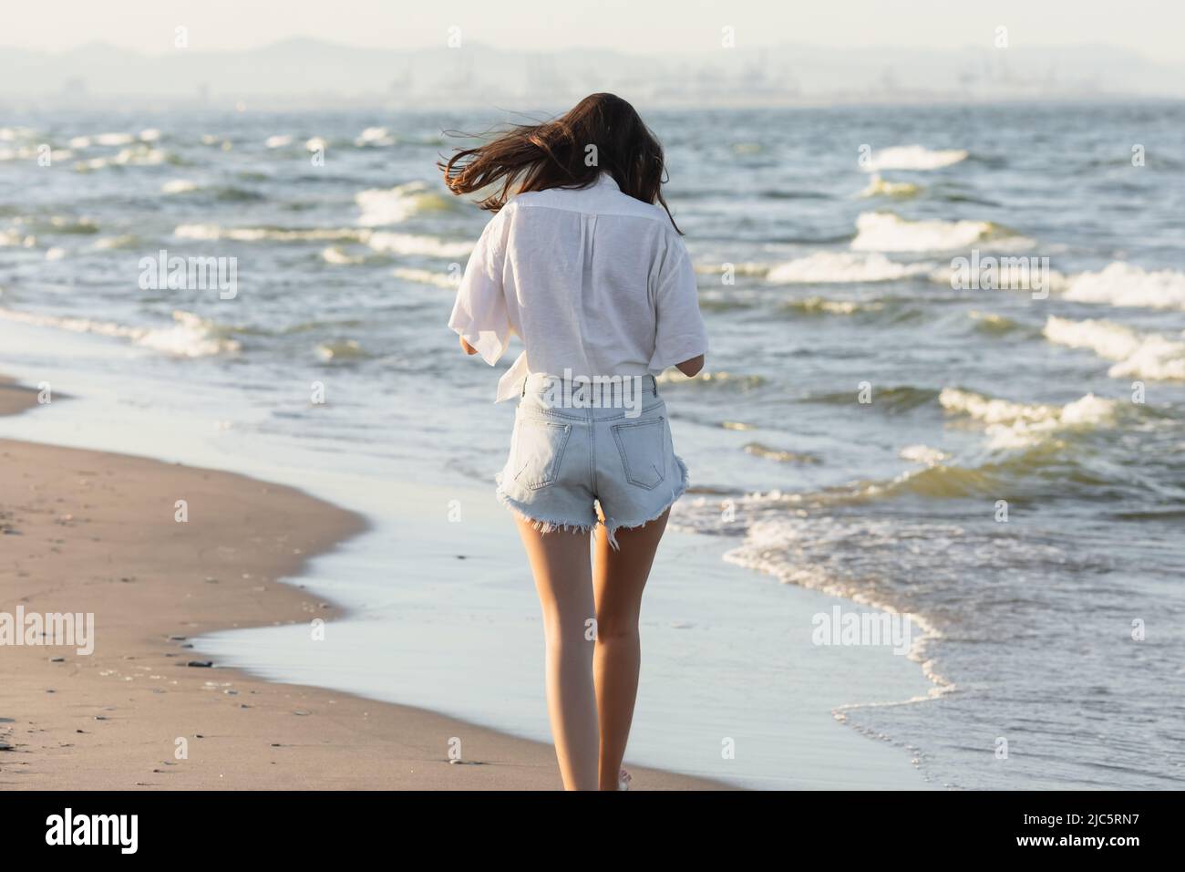 Back view of woman in white shirt and denim shorts walking near sea on ...