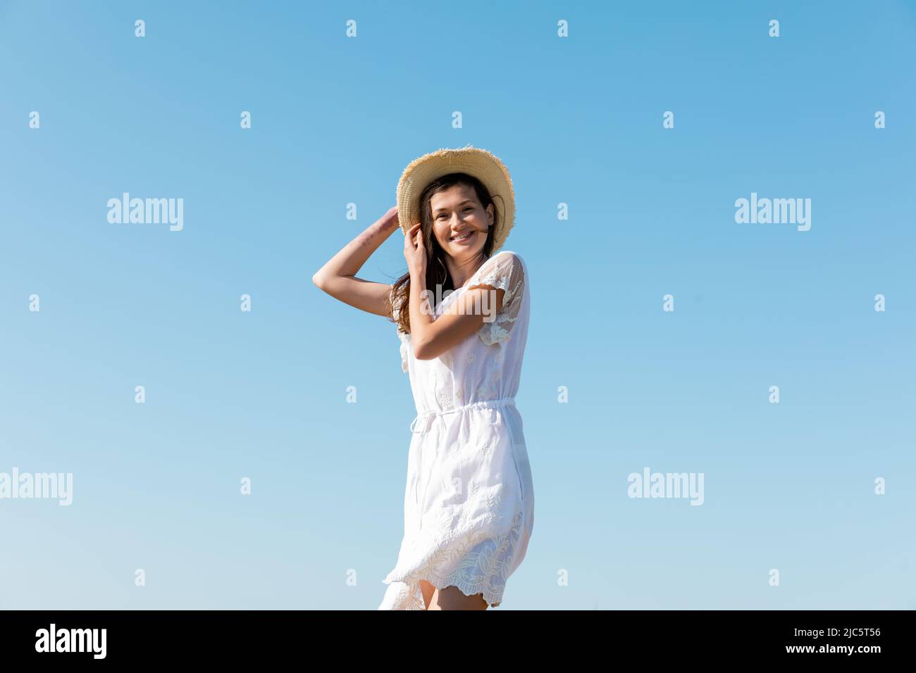 Low angle view of smiling woman in sun hat and dress looking at camera ...