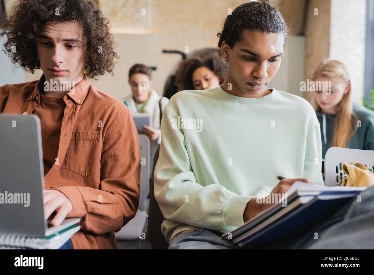 african american student writing in notebook while friend typing on ...