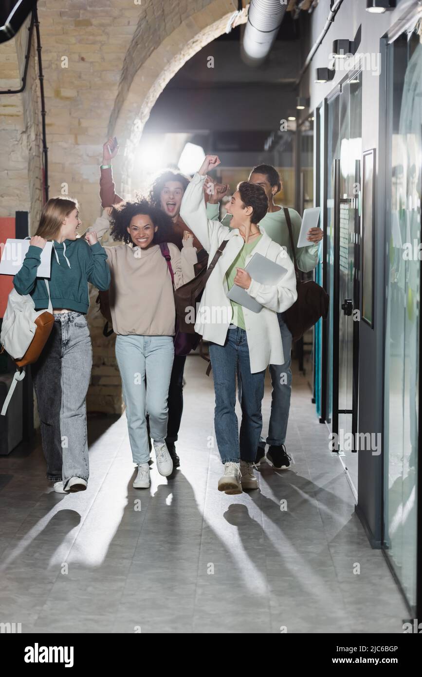 excited interracial friends walking along university hallway and ...