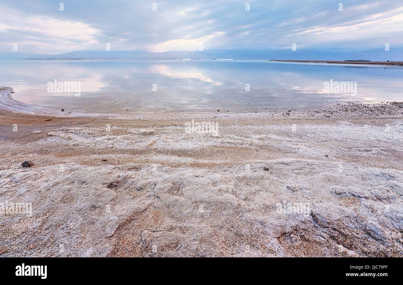 Sand and stones covered with crystalline salt on shore of Dead Sea, calm clear water near ...