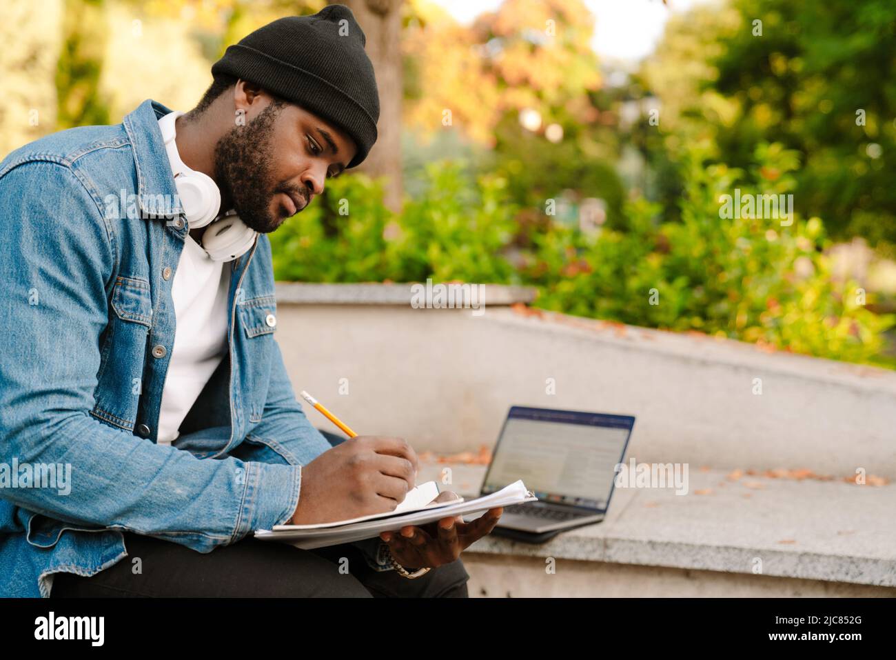 Black man working with laptop and papers while sitting on bench in park ...