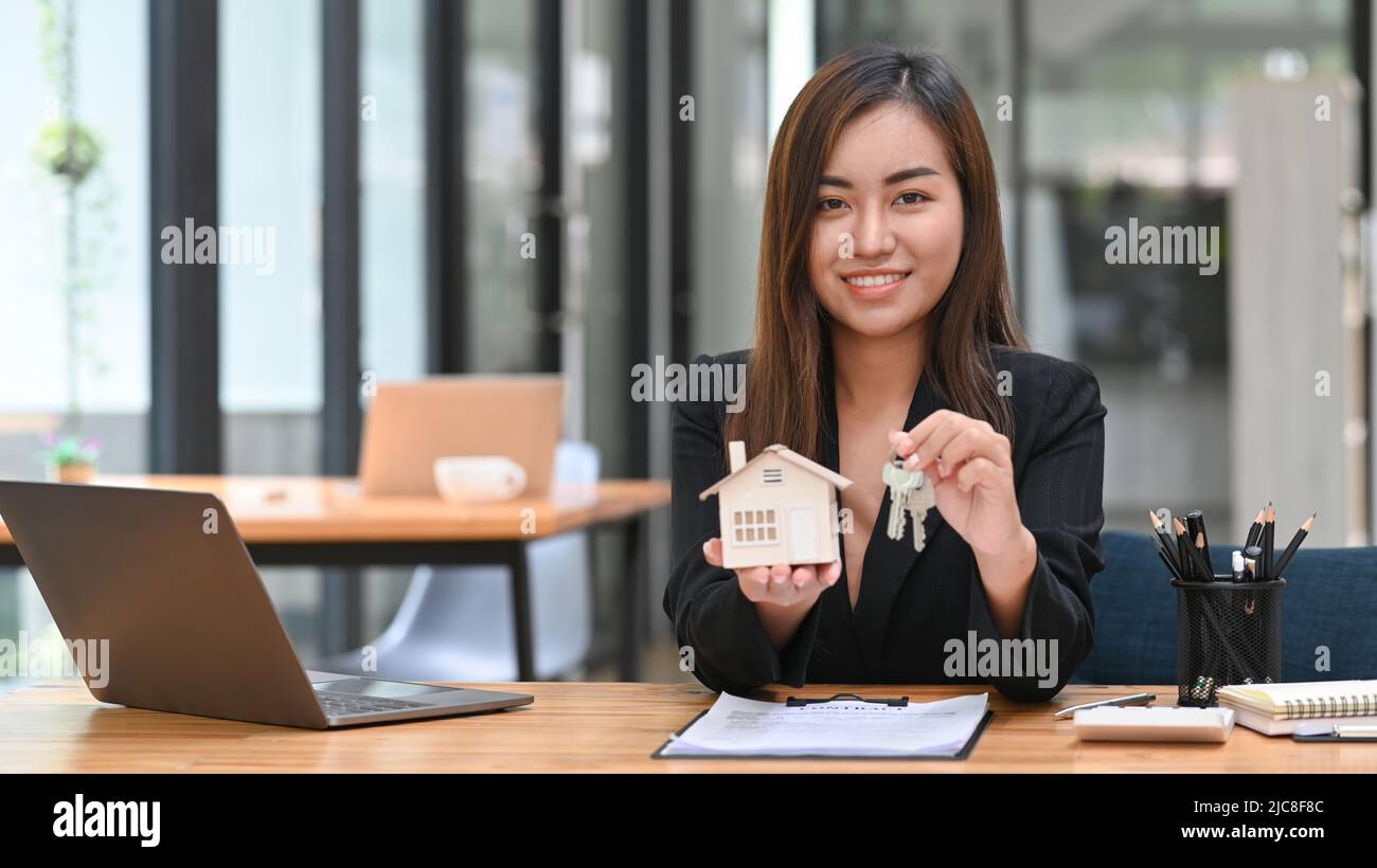 Smiling young woman agent holding small house model and keys. Real ...