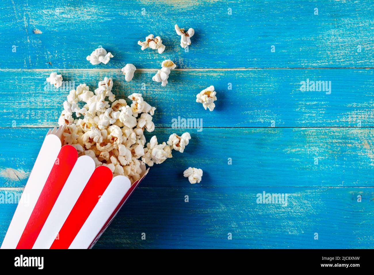 Fresh popcorn spilled from a cardboard striped cup on a blue wooden ...
