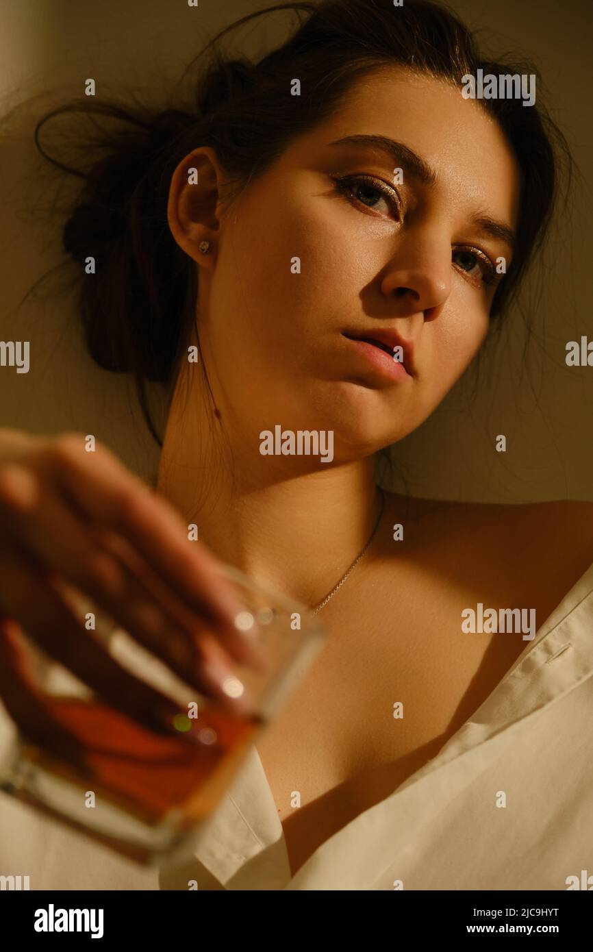 Young woman drinking alcohol from glass against sunny wall background ...